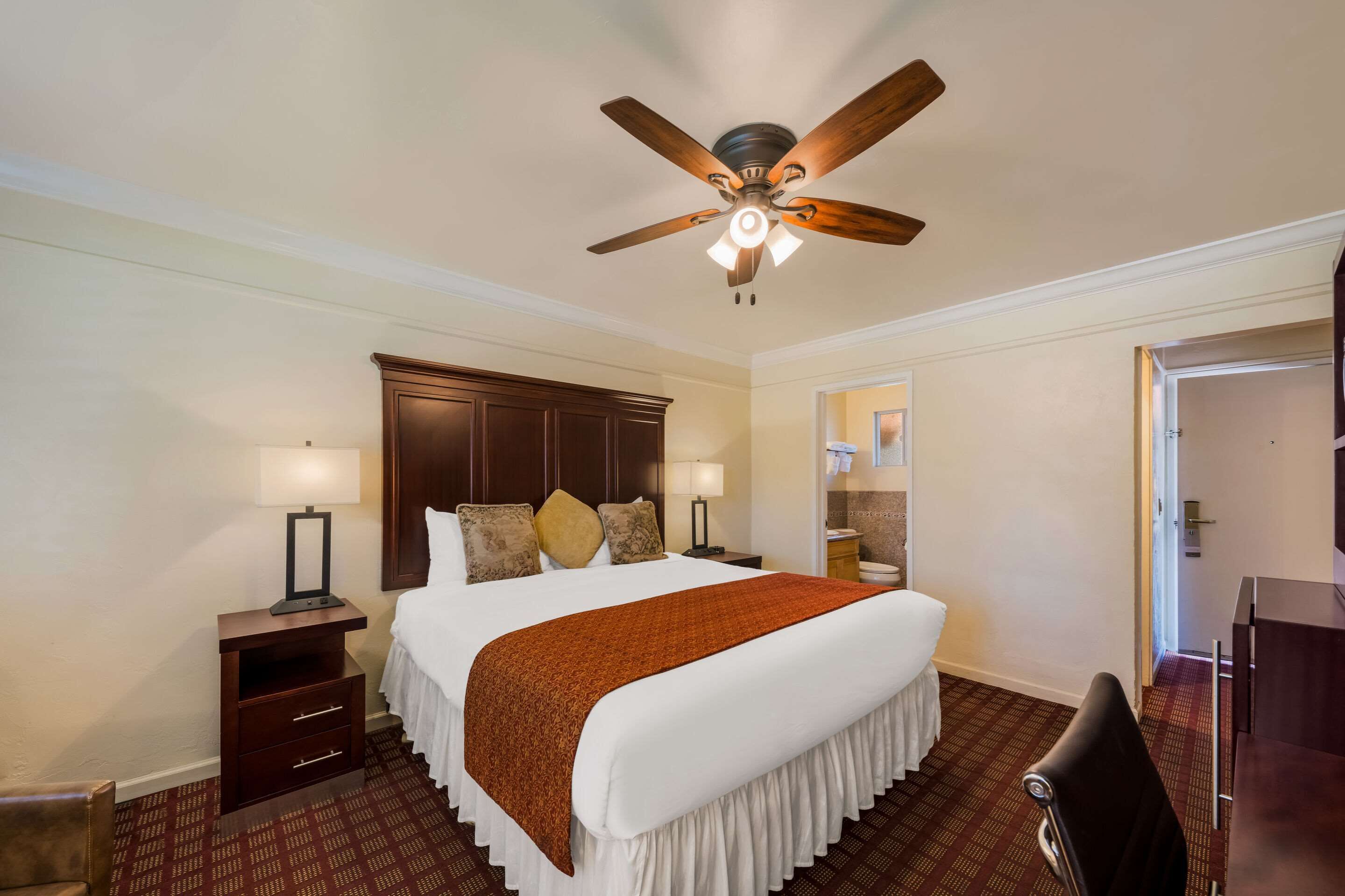 Quarto de hotel com uma cama grande, um ventilador de teto, uma mesa de cabeceira com abajur e um espelho.