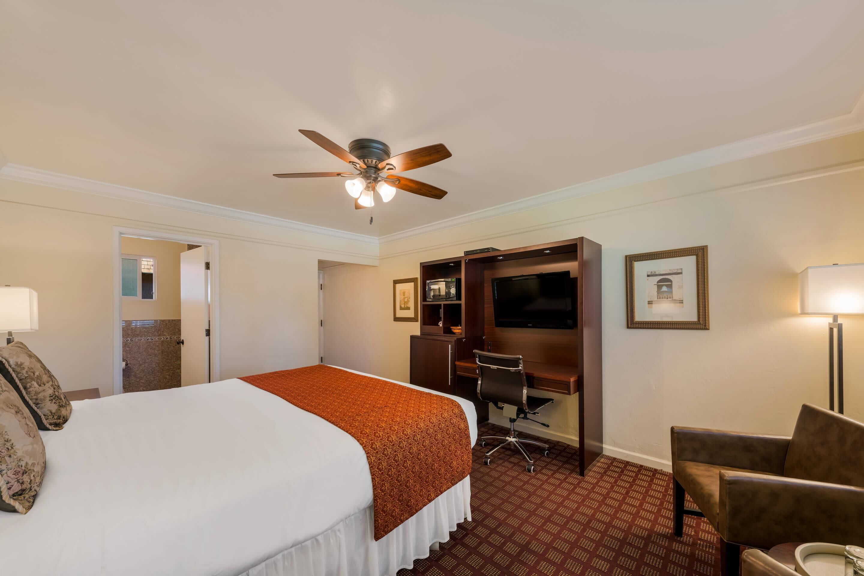 Quarto de hotel com cama, escrivaninha, cadeira, ventoinha no teto, televisão e quadros na parede.