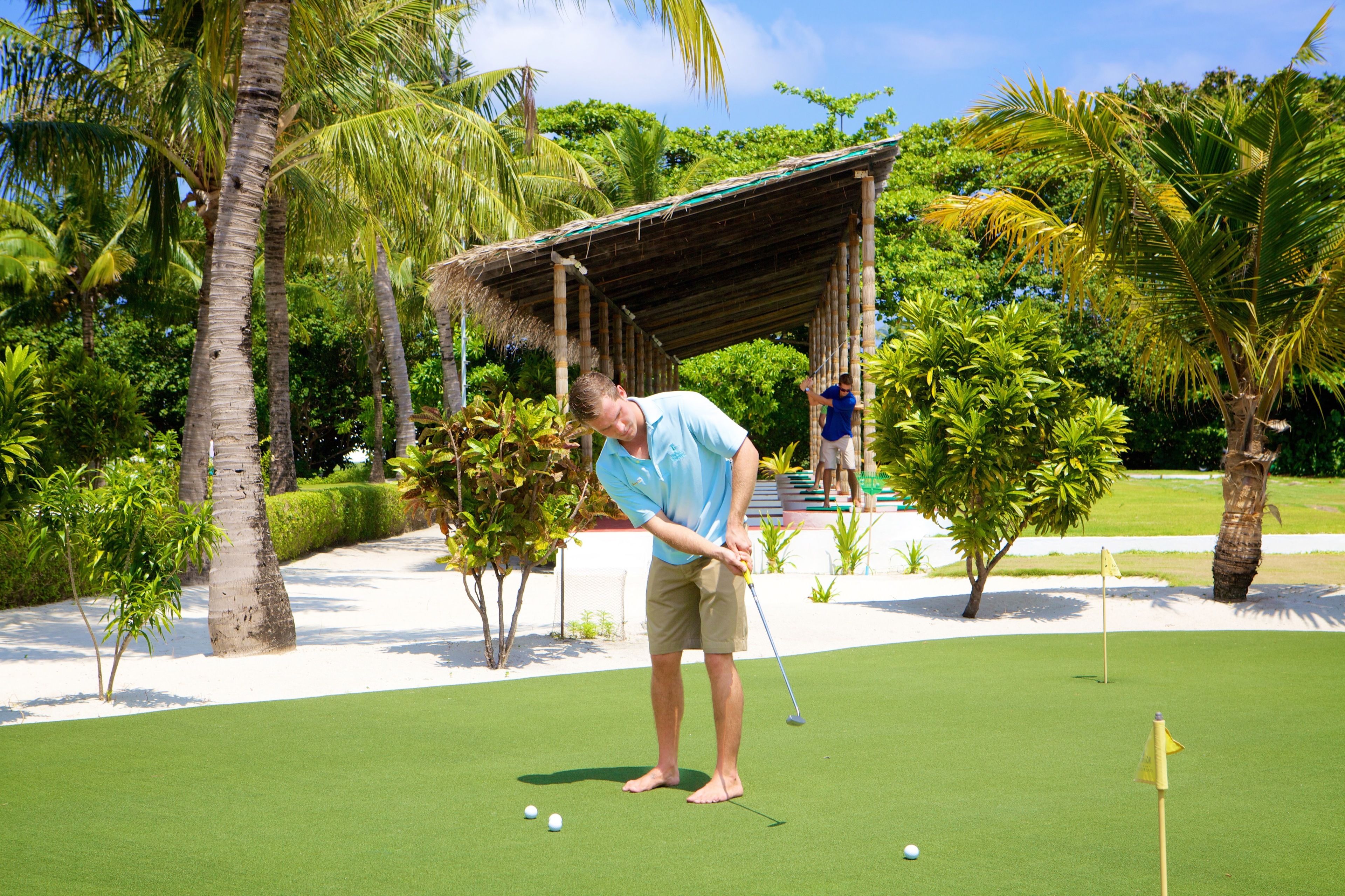 Um homem joga golfe num campo, com uma construção de telhado de palha ao fundo.