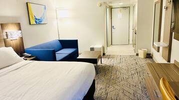 Quarto de hotel com cama, sofá azul, escrivaninha e cadeira. Há um corredor que leva a outro quarto.