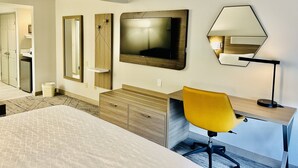 Quarto de hotel com cama, escrivaninha, cadeira, televisão, espelho e pequeno armário.
