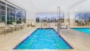 Piscina interna com água azul cristalina, rodeada por espreguiçadeiras e mesas, amplas janelas com vista para o exterior.