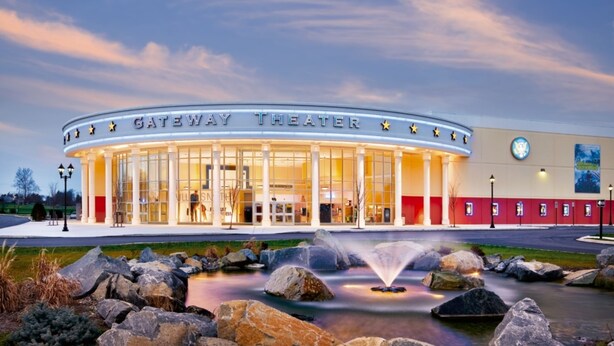 O Gateway Theater com uma entrada imponente, uma fonte em primeiro plano e uma área ajardinada com pedras.