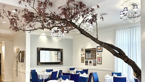 Restaurante com cadeiras azuis e toalhas de mesa brancas, um candelabro e um galho de árvore como peça central.