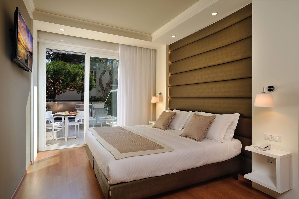 Quarto de hotel moderno com uma cama grande, uma mesinha de cabeceira com luminária e vista para o exterior por uma porta de vidro deslizante.