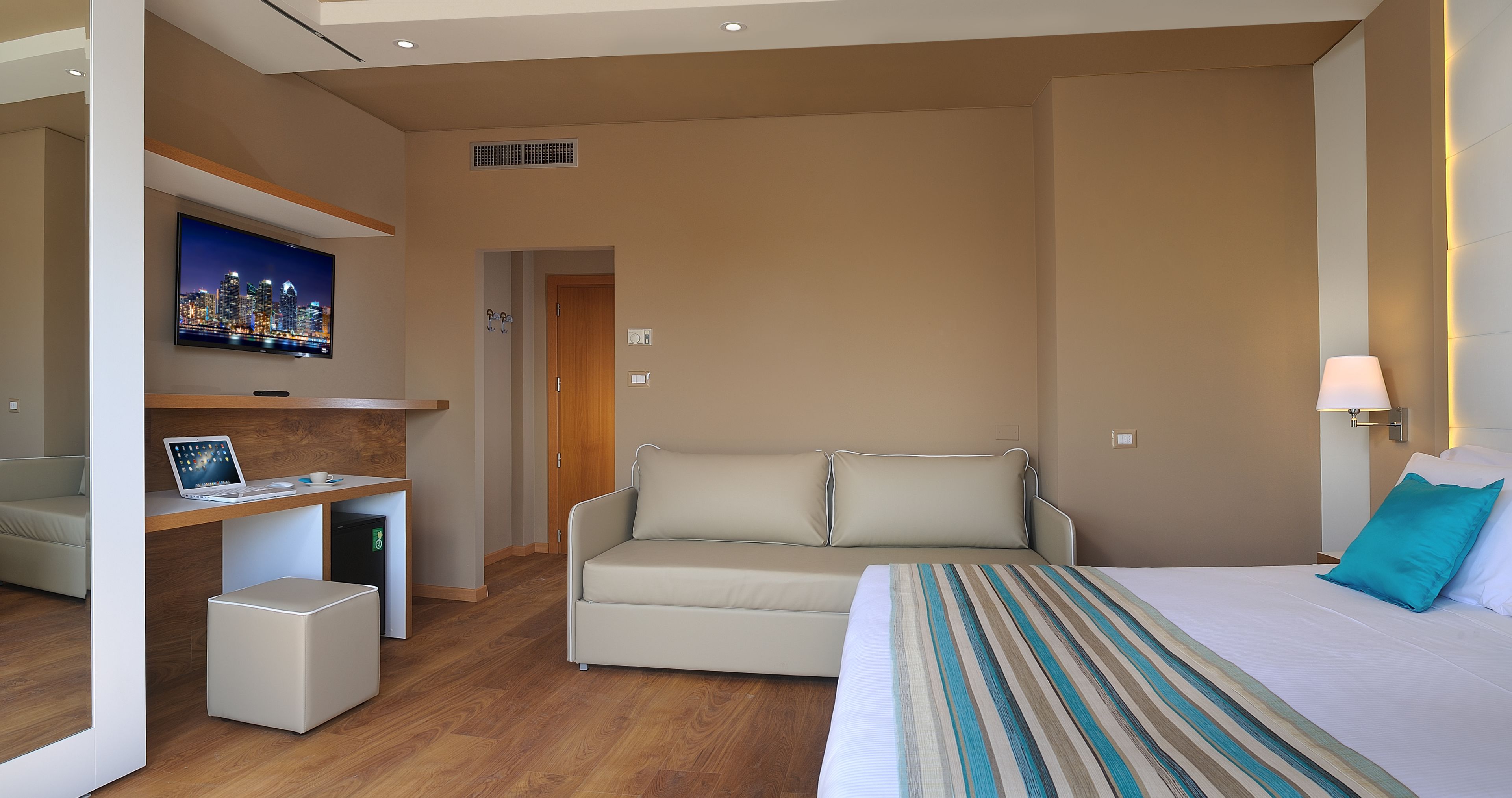 Quarto de hotel moderno com uma cama, um sofá, uma escrivaninha com um laptop, uma televisão e um pequeno armário.