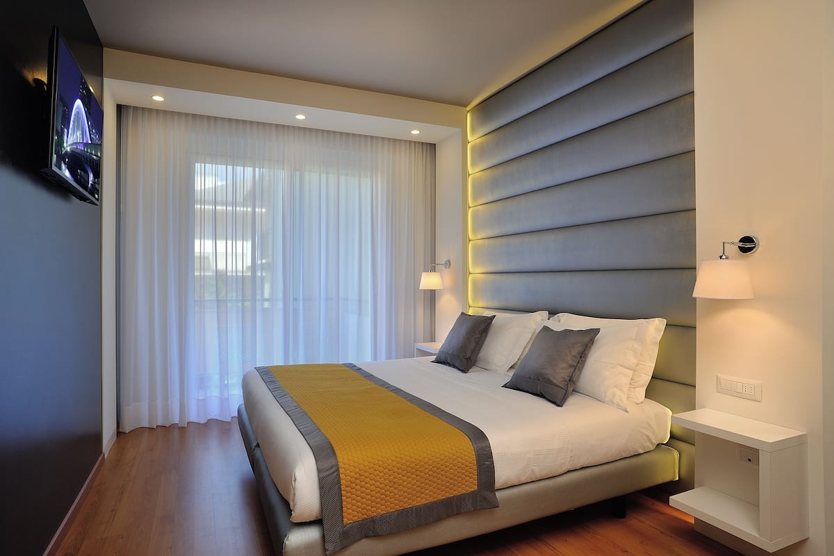 Quarto de hotel moderno com uma cama grande, televisão fixada na parede e uma janela com cortinas leves.