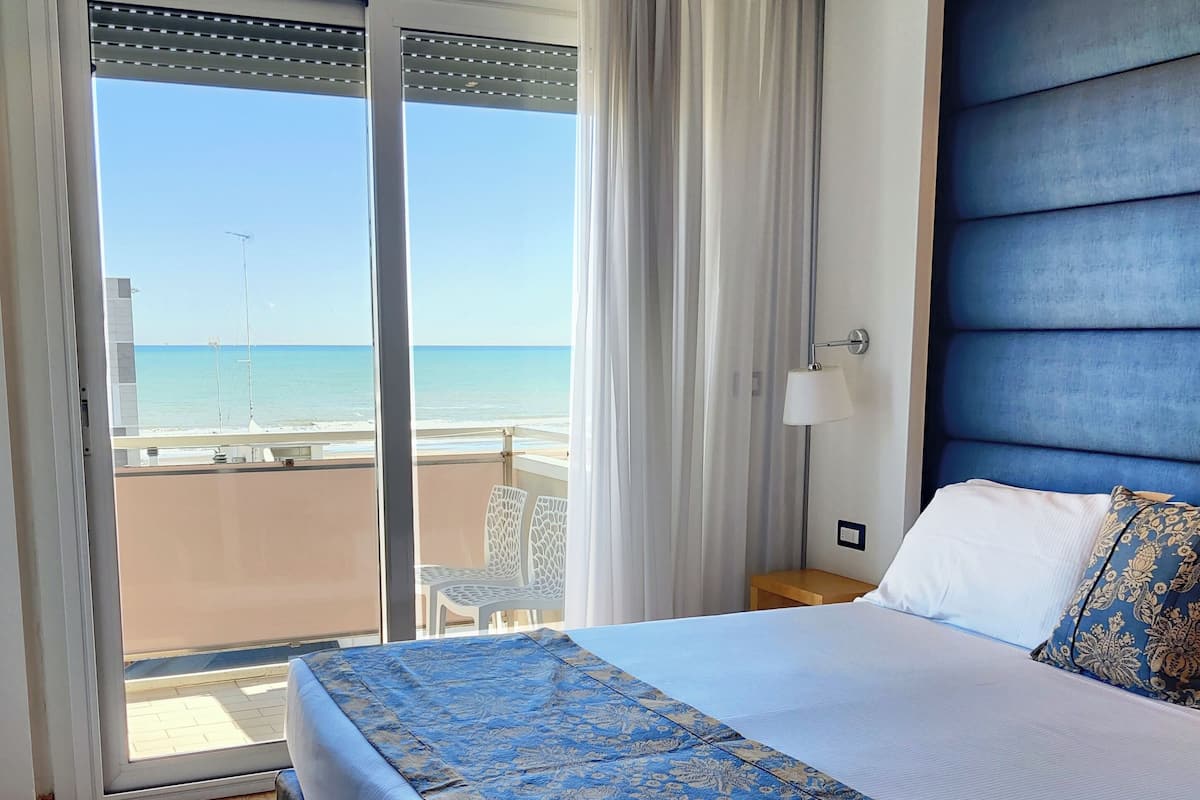 Quarto de hotel com uma cama grande, vista para a praia e porta de vidro que se abre para a varanda.