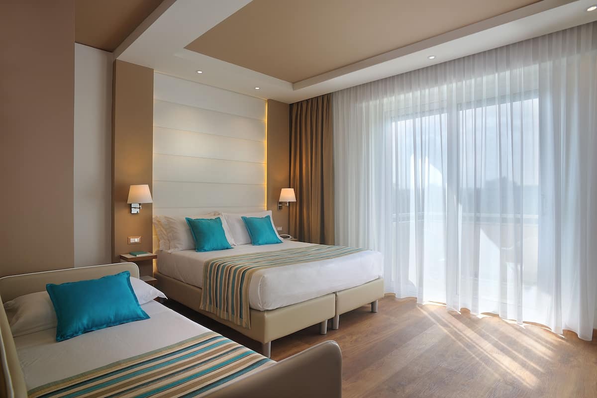 Quarto de hotel com uma cama grande, duas mesas de cabeceira e vista para o exterior através de cortinas leves.