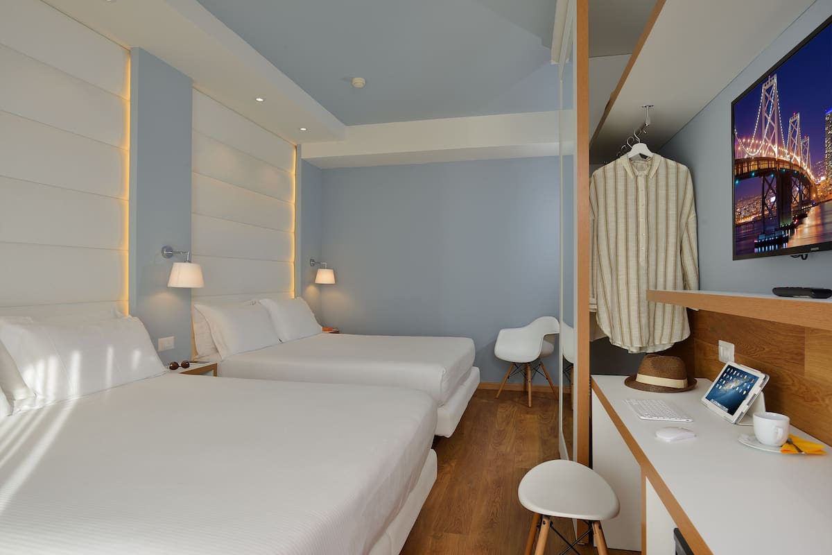 Quarto de hotel com duas camas, uma escrivaninha com computador, uma cadeira, uma televisão e um quadro de uma ponte.