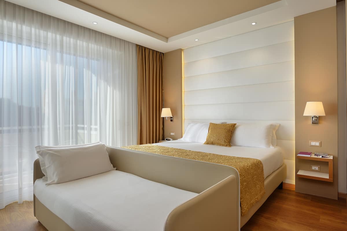 Quarto de hotel moderno com uma cama grande, um sofá e uma mesa de cabeceira com abajur.