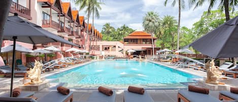 Uma área de piscina com espreguiçadeiras e guarda-sóis, cercada por palmeiras e um edifício com telhado vermelho.