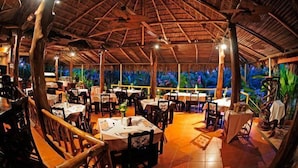 Restaurante interno com telhado de palha, mesas e cadeiras de madeira.