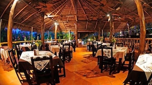 Restaurante interno com mesas e cadeiras de madeira, teto de palha e vista do céu noturno.