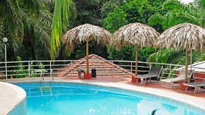 Piscina com espreguiçadeiras e guarda-sóis de palha, rodeada por vegetação exuberante.