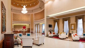 Um hall amplo com teto alto, lustres elegantes e uma área de estar espaçosa com sofás e poltronas brancas.