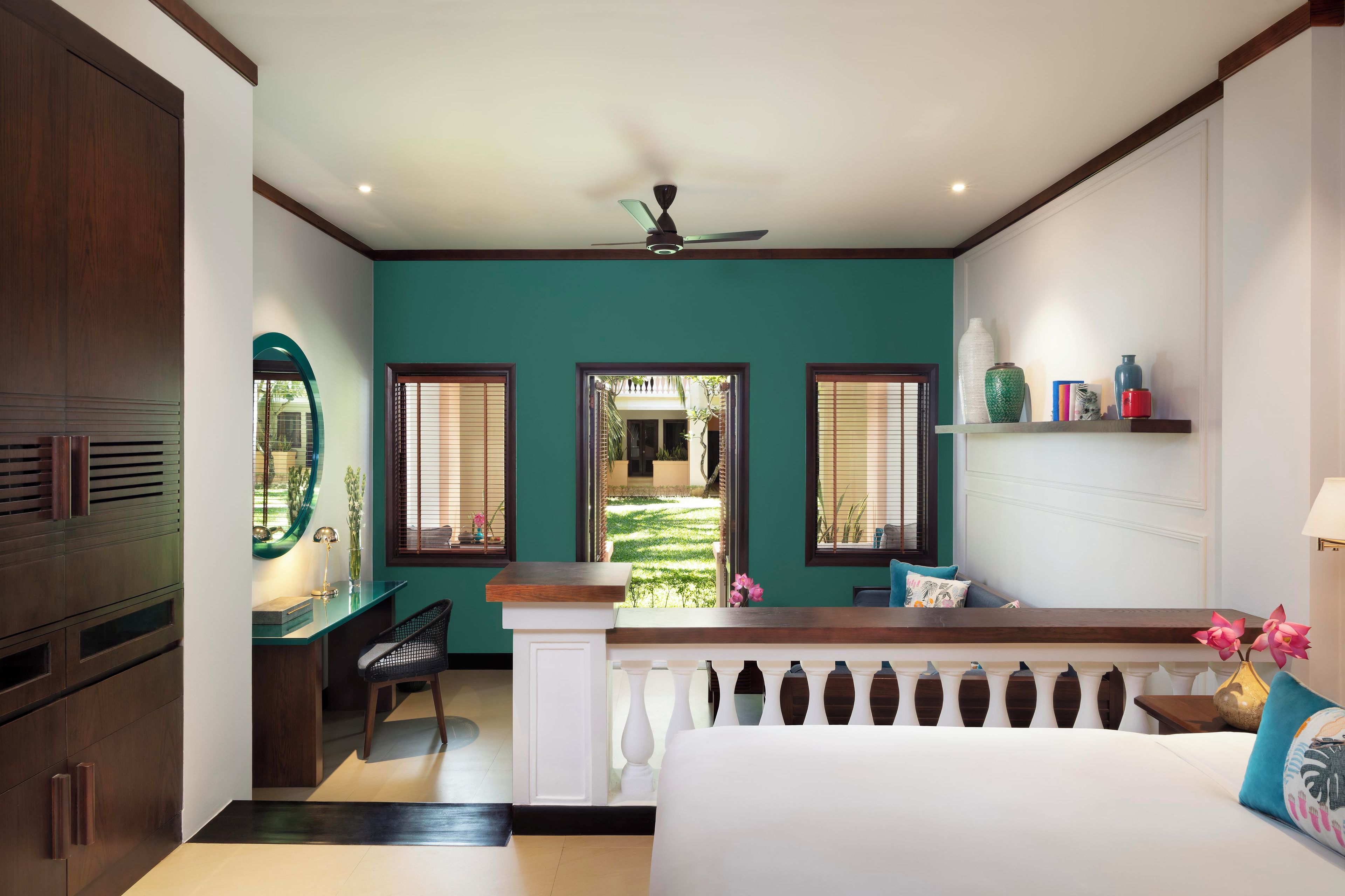 Quarto com cama, escrivaninha, cadeira, ventilador de teto e vista para o exterior.