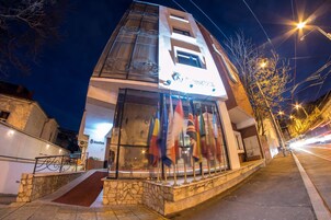 Um edifício moderno com fachada de vidro, entrada com tapete vermelho e várias bandeiras hasteadas na parte externa.