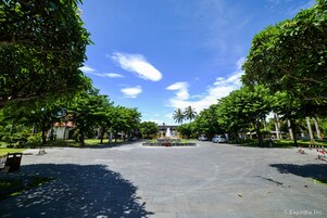 Um pátio amplo com uma fonte central, rodeado por árvores verdes exuberantes e um céu azul límpido.