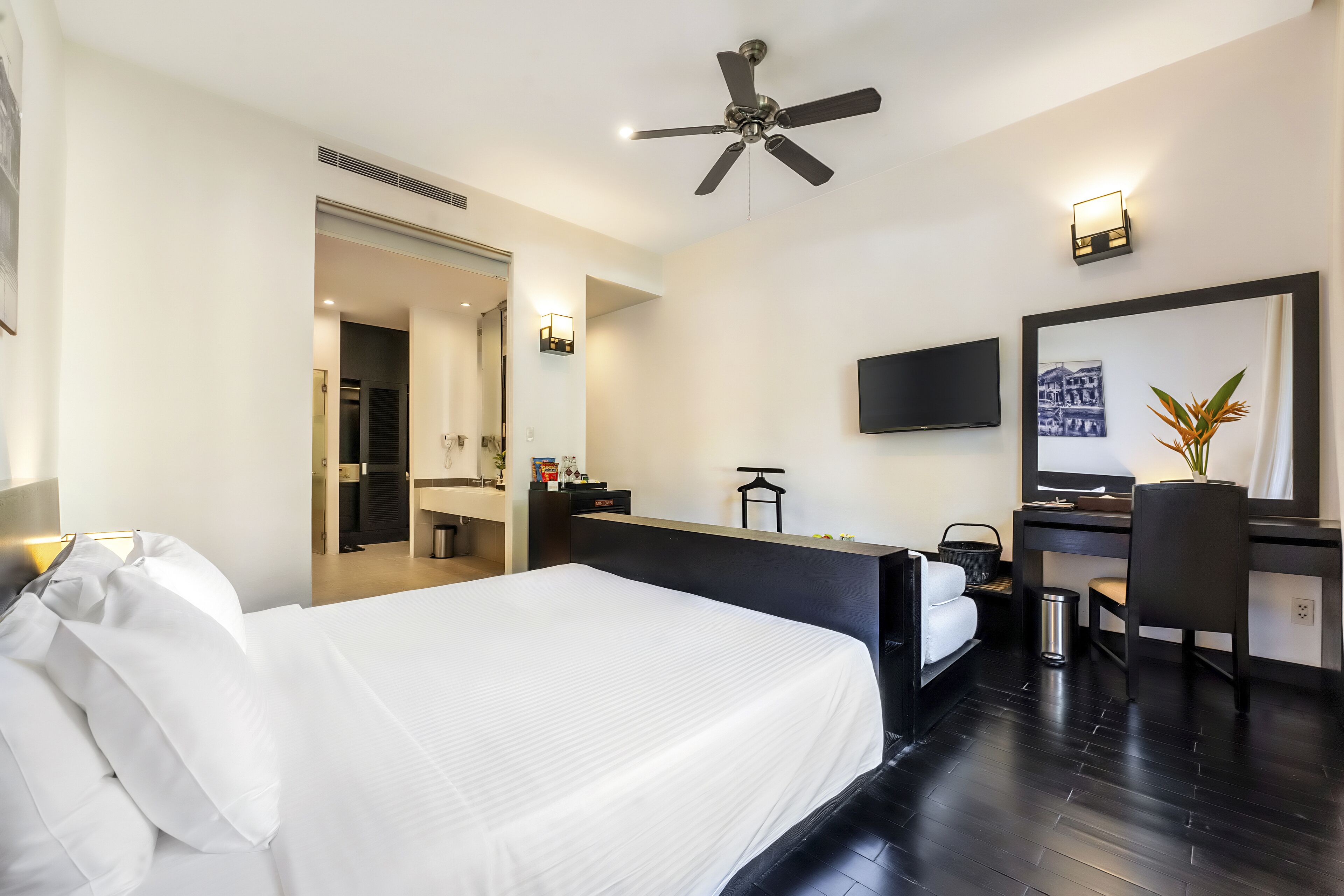 Quarto de hotel com cama, escrivaninha, cadeira, televisão e ventoinha no teto.
