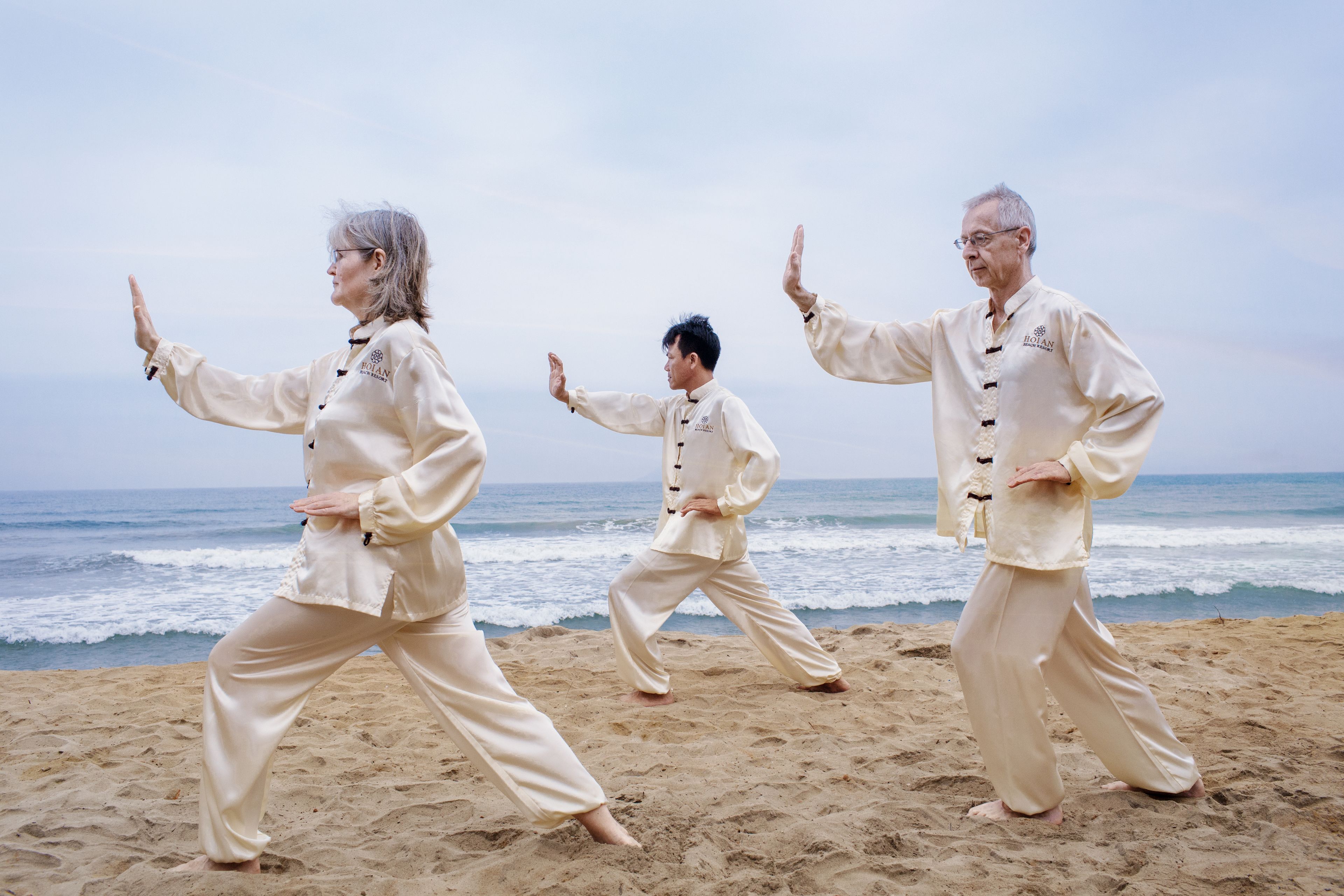 Três pessoas praticam Tai Chi numa praia, com o oceano ao fundo.