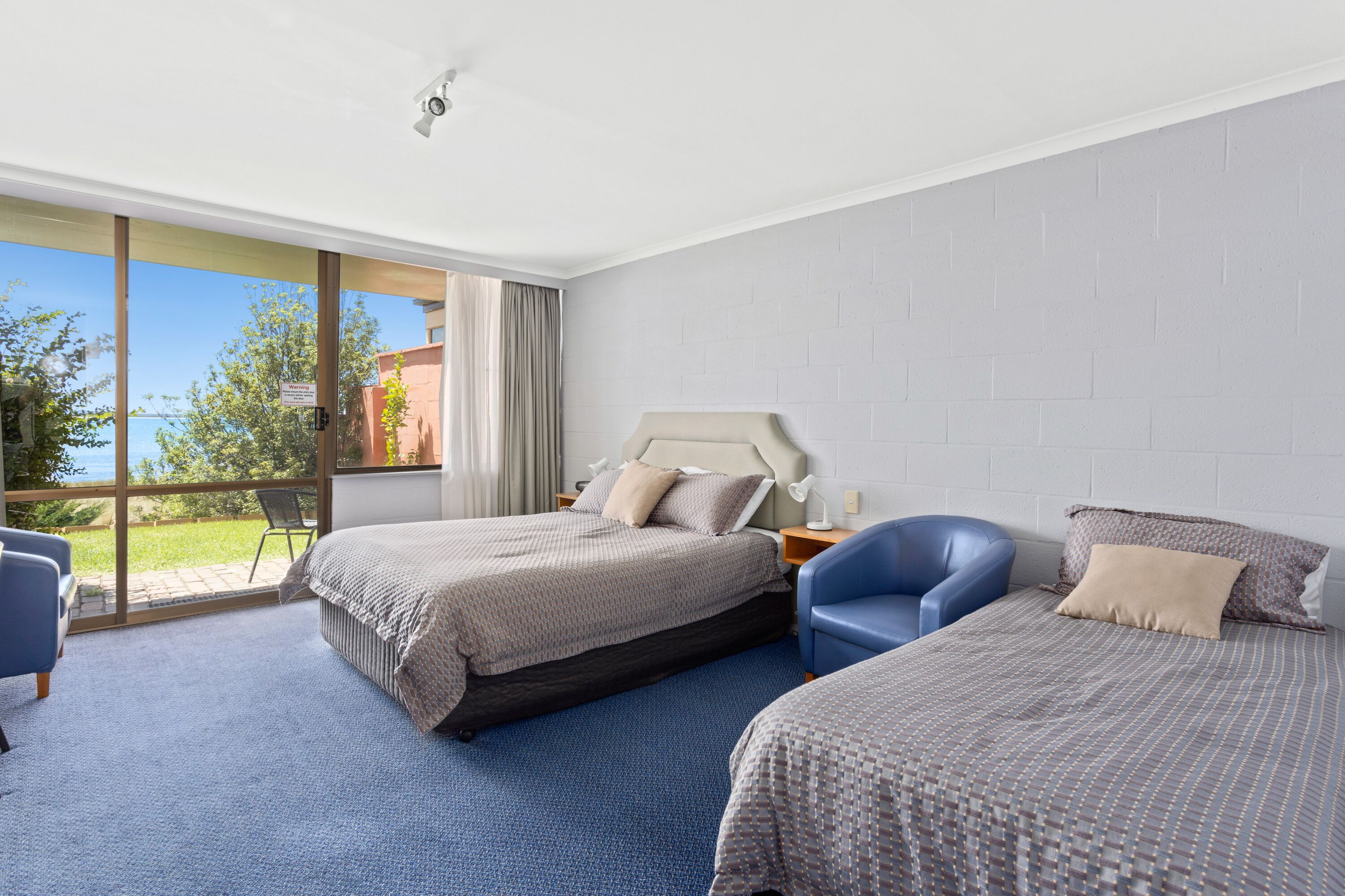 Quarto de hotel com duas camas, uma cadeira azul e uma janela grande com vista para o exterior.