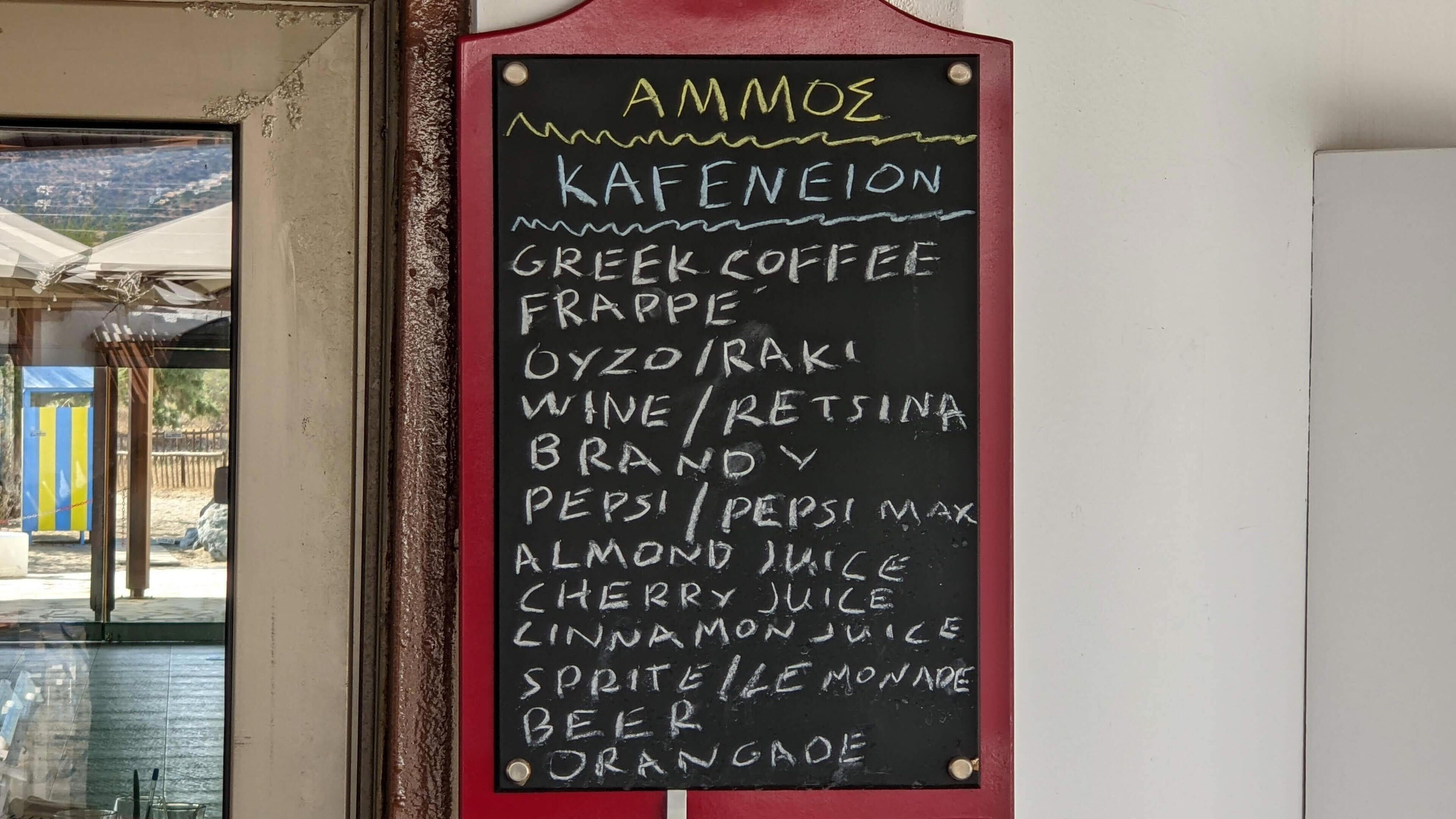 Quadro-negro com cardápio listando pratos e bebidas como café grego, frappé e diversos sucos.