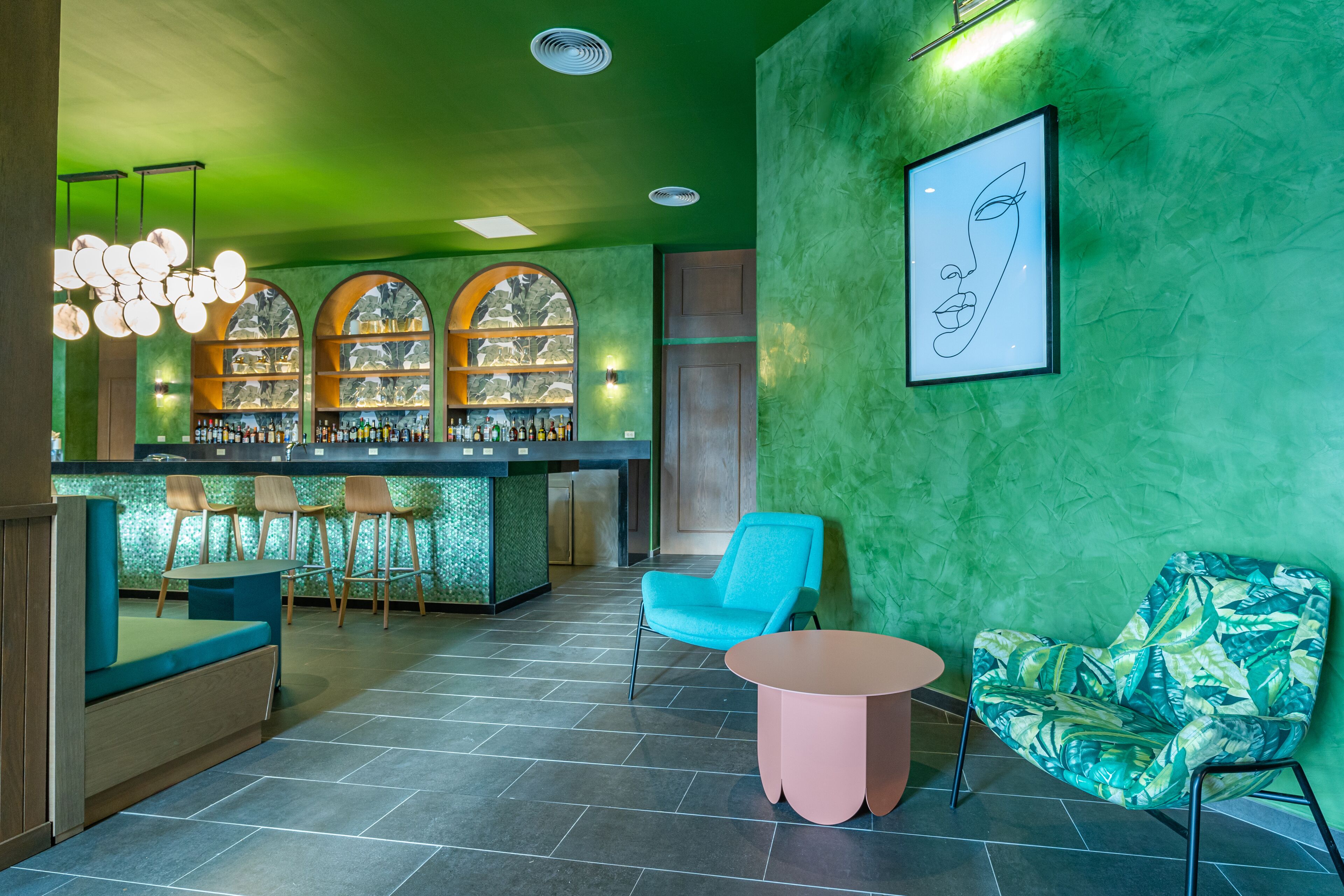 Uma área de bar com parede verde, uma obra de arte emoldurada e um espaço de estar com uma mesa redonda e duas cadeiras.