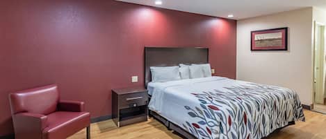 Quarto de hotel com cama, uma cadeira vermelha, mesa de cabeceira e um quadro pendurado na parede.