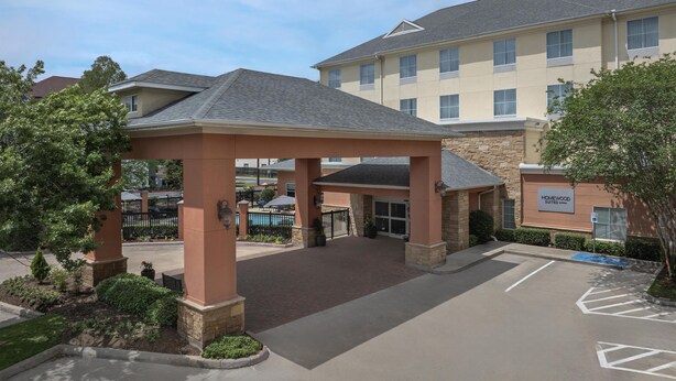Entrada de hotel com área de espera coberta, estacionamento e um edifício com uma placa que diz 'Homewood Suites'.