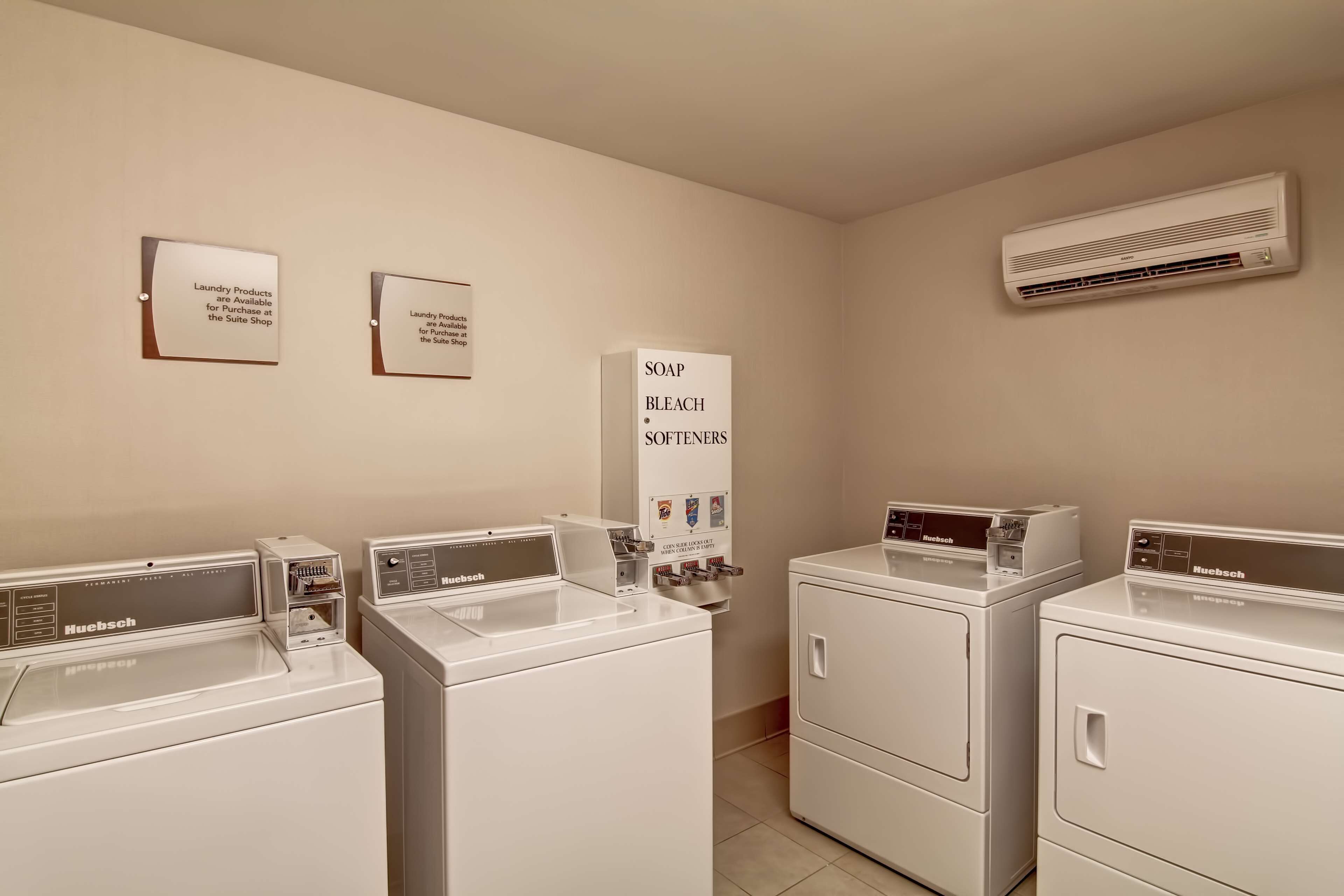 Uma lavanderia com três máquinas de lavar brancas, um letreiro com os produtos disponíveis e um aparelho de ar condicionado na parede.