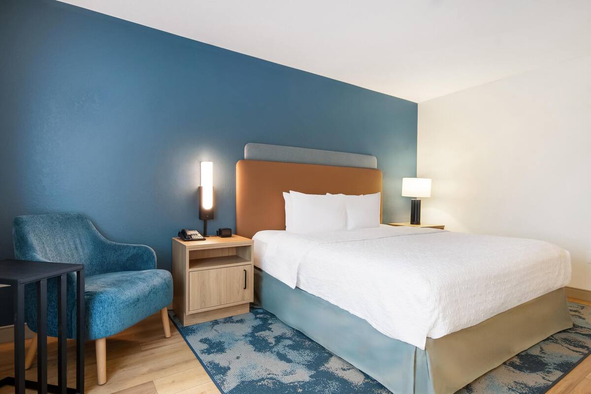 Quarto de hotel com cama, poltrona, mesa de cabeceira e escrivaninha.