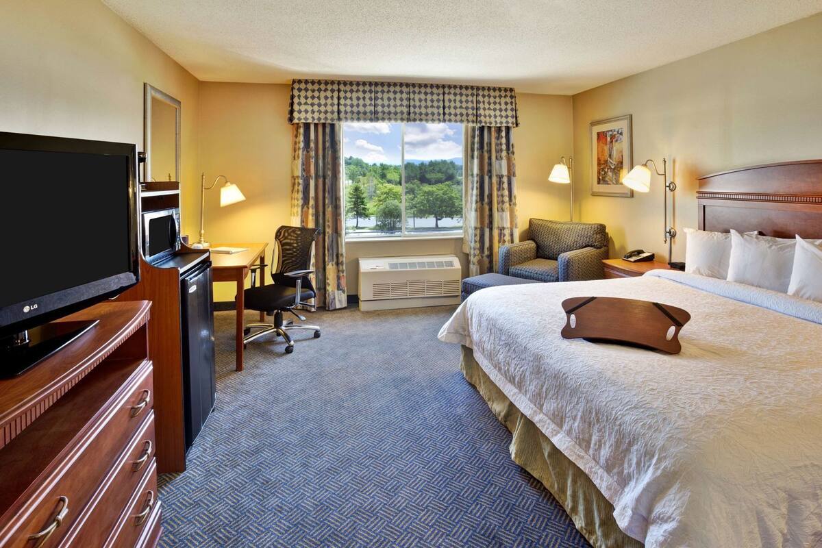 Quarto de hotel com uma cama, escrivaninha, televisão e janela com cortinas.