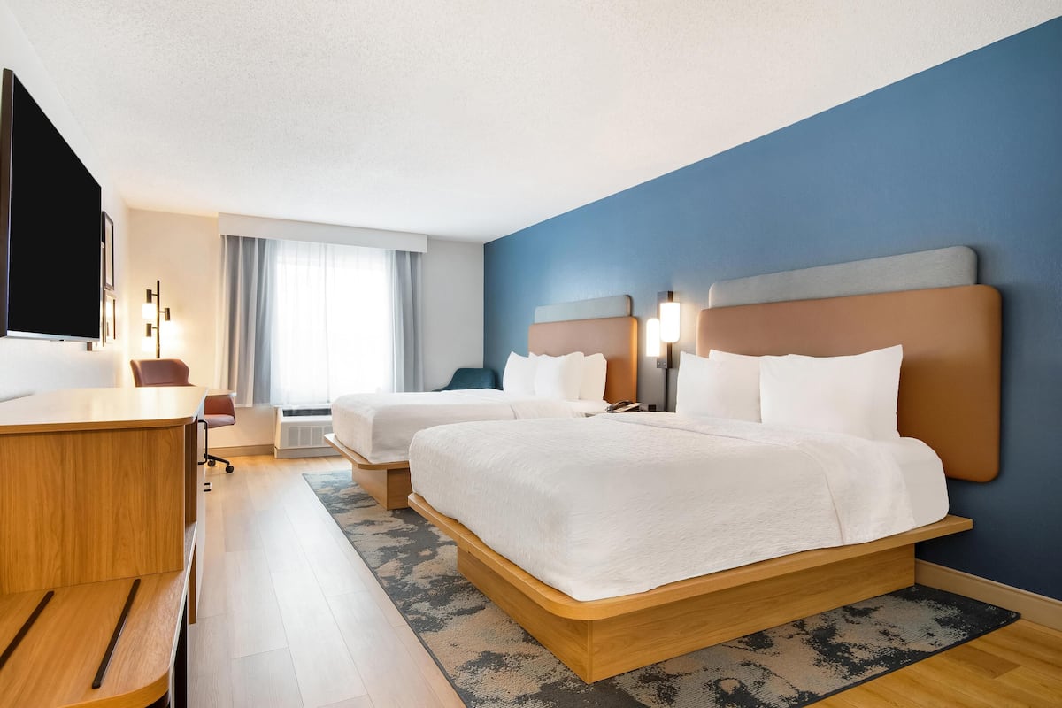 Quarto de hotel com duas camas, uma escrivaninha, televisão de tela plana e uma janela grande com cortinas.
