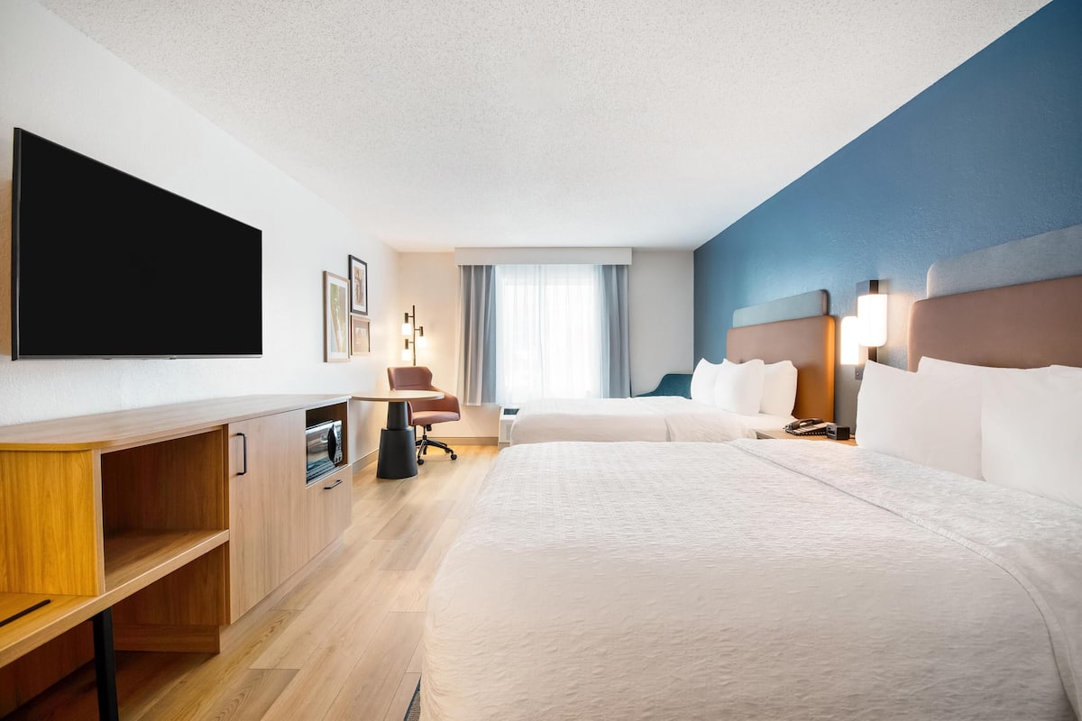 Quarto de hotel com cama grande, escrivaninha, TV de tela plana e janela com cortinas.