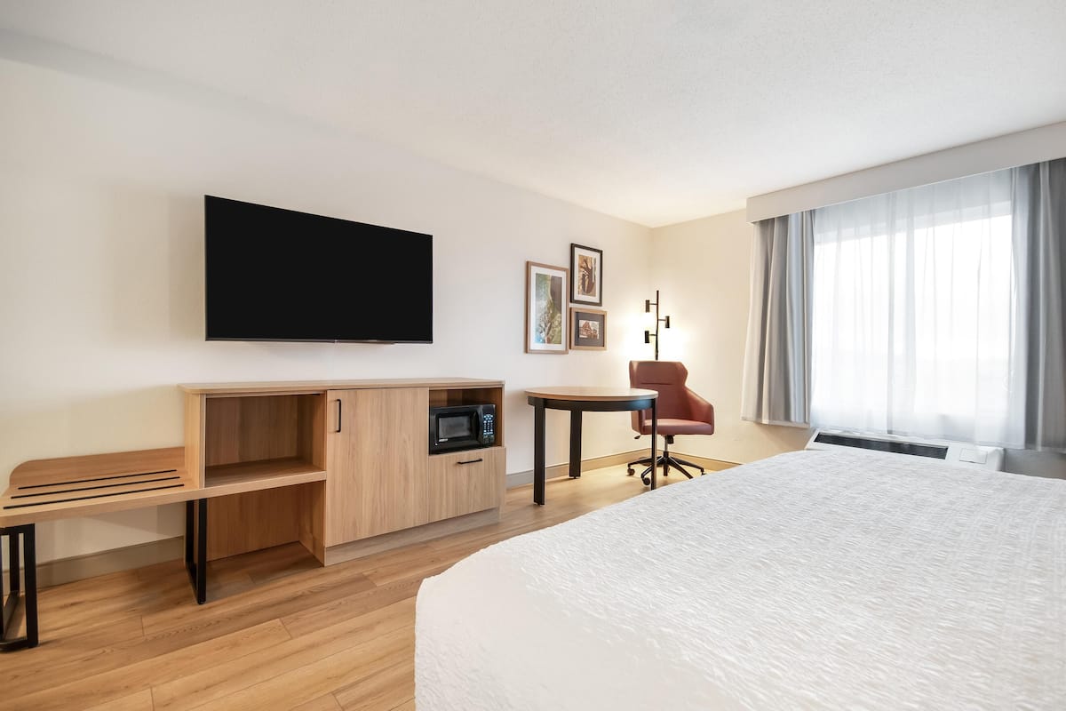 Quarto de hotel com cama, secretária com micro-ondas, cadeira, televisão montada na parede e janela com cortinas.