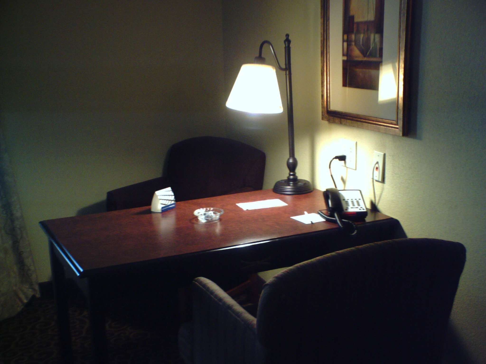 Quarto de hotel com uma escrivaninha de madeira, uma lâmpada, um telefone e uma cadeira.