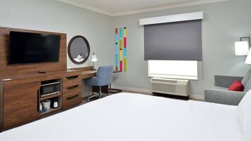 Quarto de hotel com cama, escrivaninha com cadeira, televisão de tela plana, espelho, janela com persianas e ar-condicionado fixado na parede.