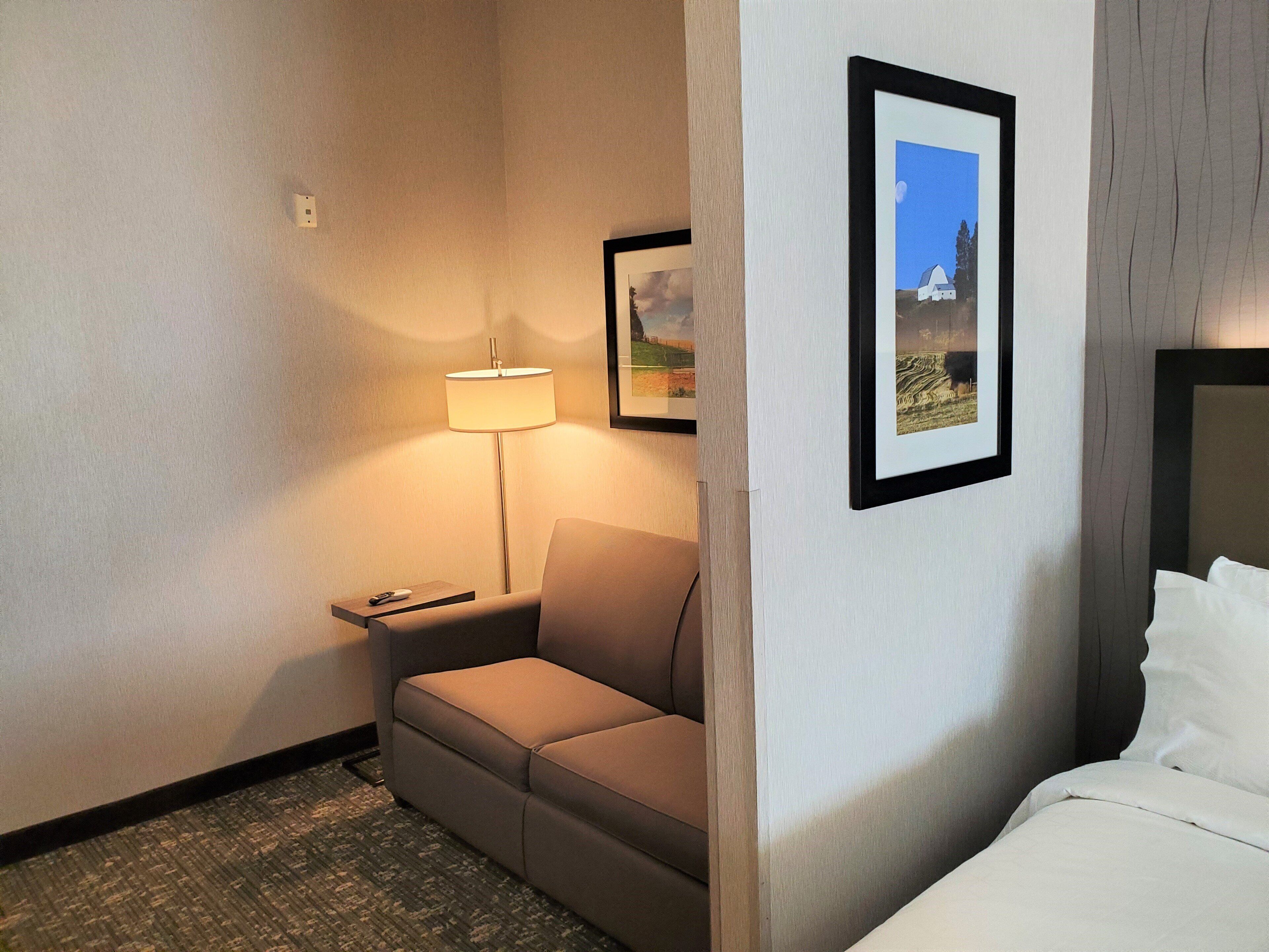 Quarto de hotel com cama, sofá, luminária e quadros na parede.
