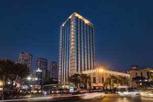 Um edifício alto com design moderno, situado em ambiente urbano com outros edifícios e uma rua movimentada.