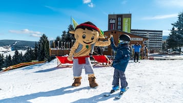 Um mascote e uma pessoa numa pista de esqui, com um hotel e árvores ao fundo.