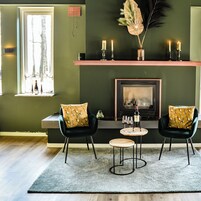 Uma sala de estar aconchegante com dois poltronas verde-escuro, uma mesinha redonda e uma lareira.