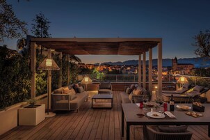 Um pátio ao ar livre com assentos, uma mesa posta para refeições e vista para uma cidade e montanhas ao fundo.