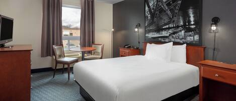 Quarto de hotel com cama, escrivaninha, cadeira, luminária e uma janela grande com cortinas.