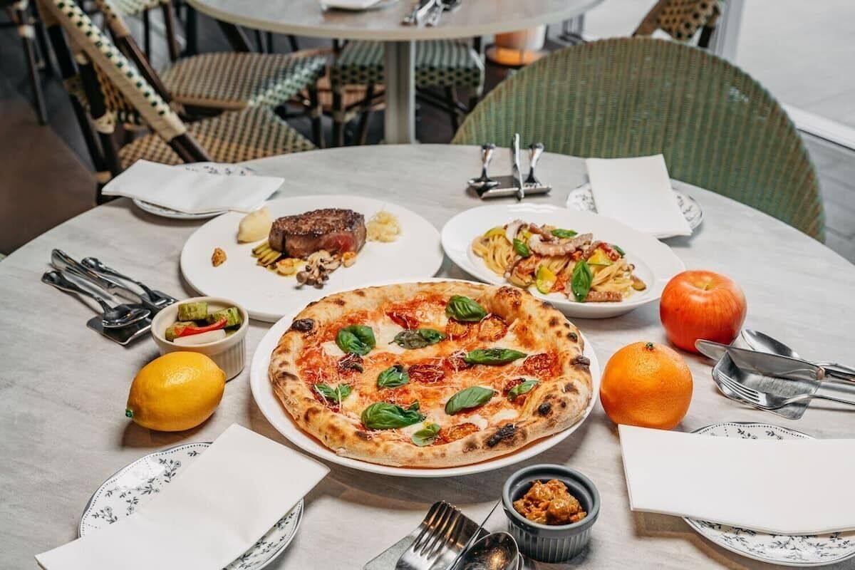 Uma mesa de jantar com diversos pratos, incluindo pizza, massa e bife, rodeada por cadeiras e utensílios de refeição.