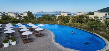 Piscina com espreguiçadeiras e guarda-sóis, rodeada por vegetação e edifícios com vista para o mar.