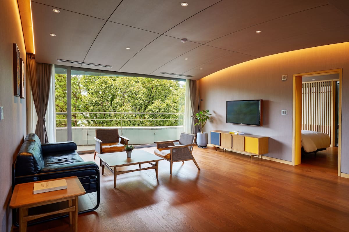 Uma sala de estar moderna com sofá, mesa de centro e cadeiras, uma televisão e uma janela grande com vista para árvores.