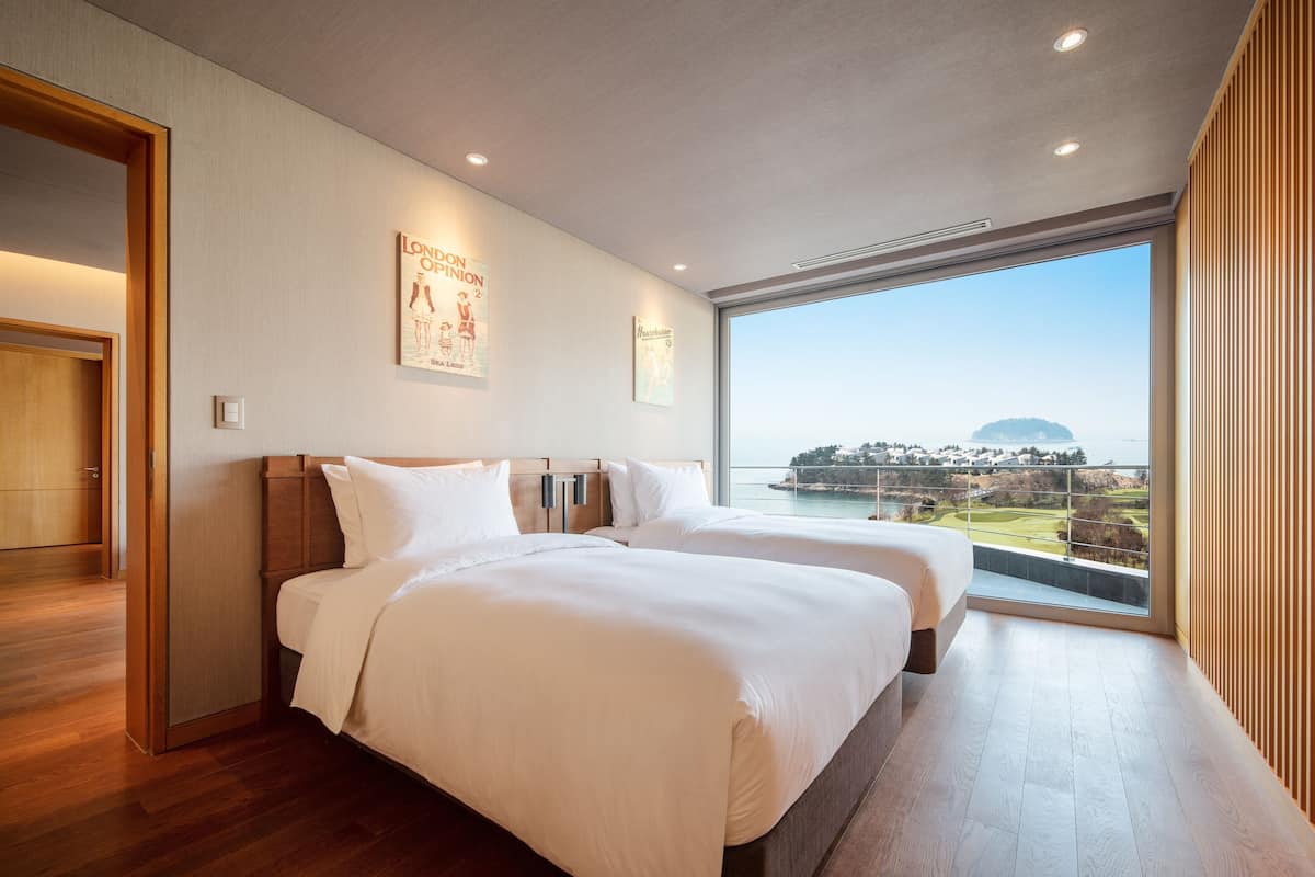 Quarto de hotel moderno com duas camas, uma janela grande com vista para uma paisagem costeira e piso de madeira.