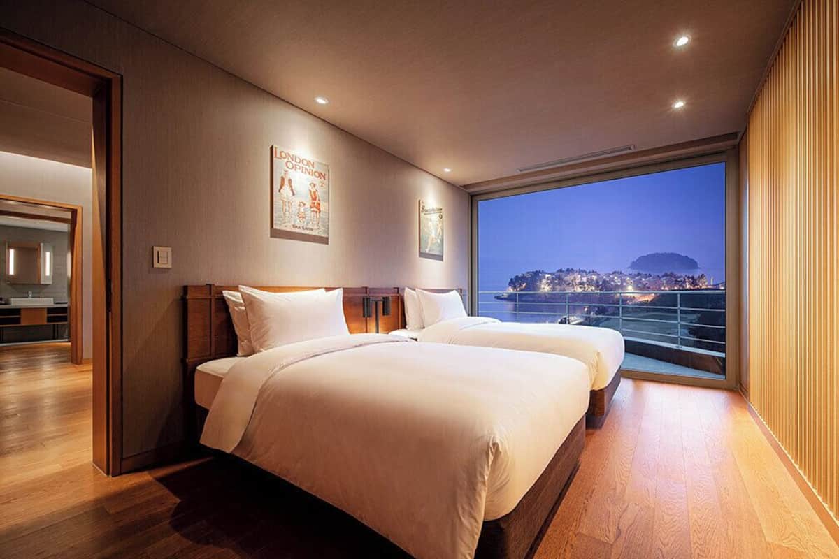 Quarto de hotel moderno com duas camas, uma janela grande com vista para a cidade e um cartaz emoldurado na parede.