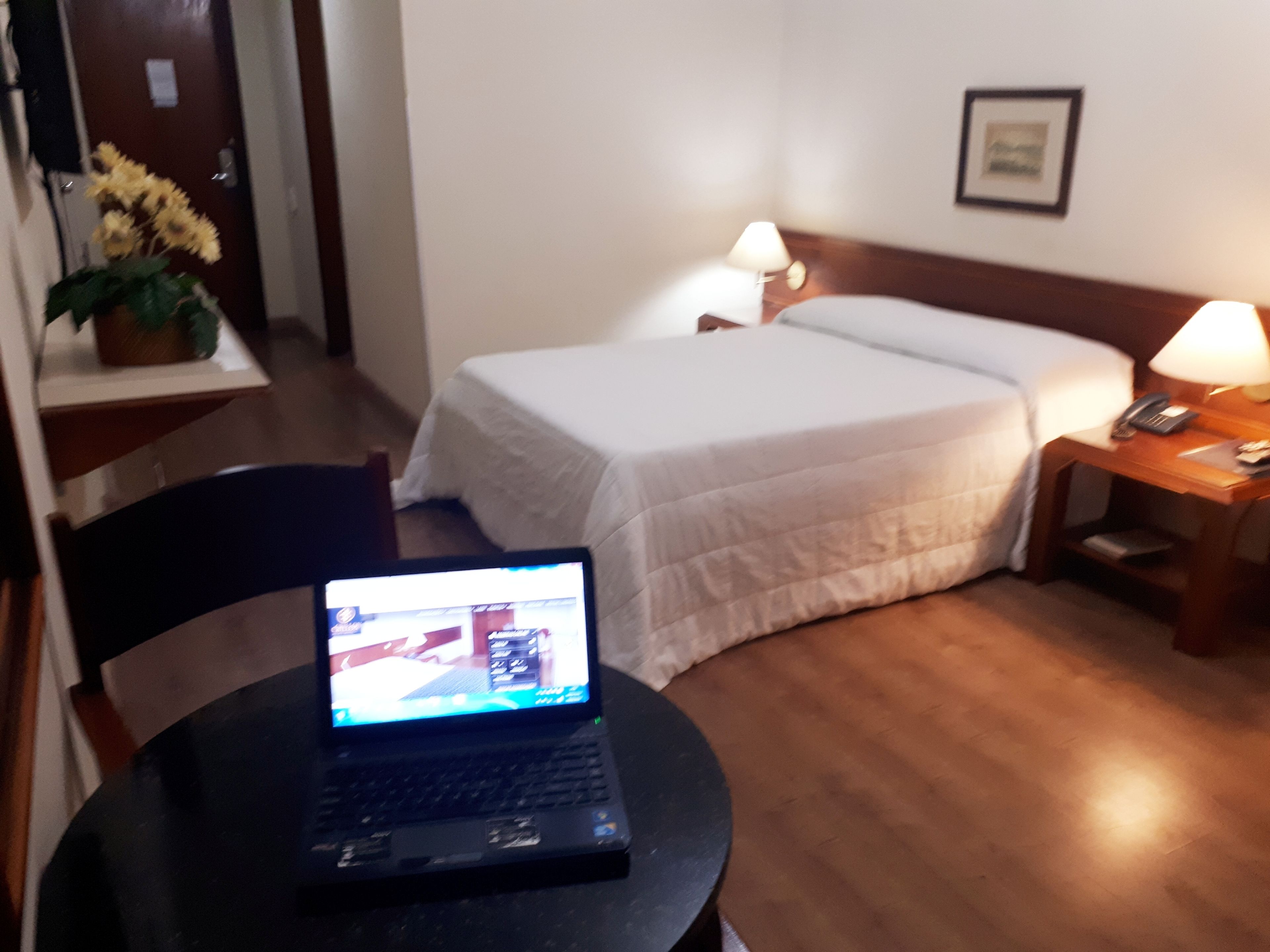 Quarto de hotel com uma cama, abajures, uma escrivaninha com um laptop e uma mesinha.